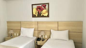 Quarto de hotel com duas camas, cada uma com colcha e travesseiros brancos, cabeceira de madeira, mesas de cabeceira com abajures e um quadro de flores pendurado na parede.