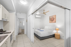 Quarto de hotel compacto com cozinha compacta, uma cama com colchão cinza, mesa de cabeceira com abajur e ventoinha no teto.