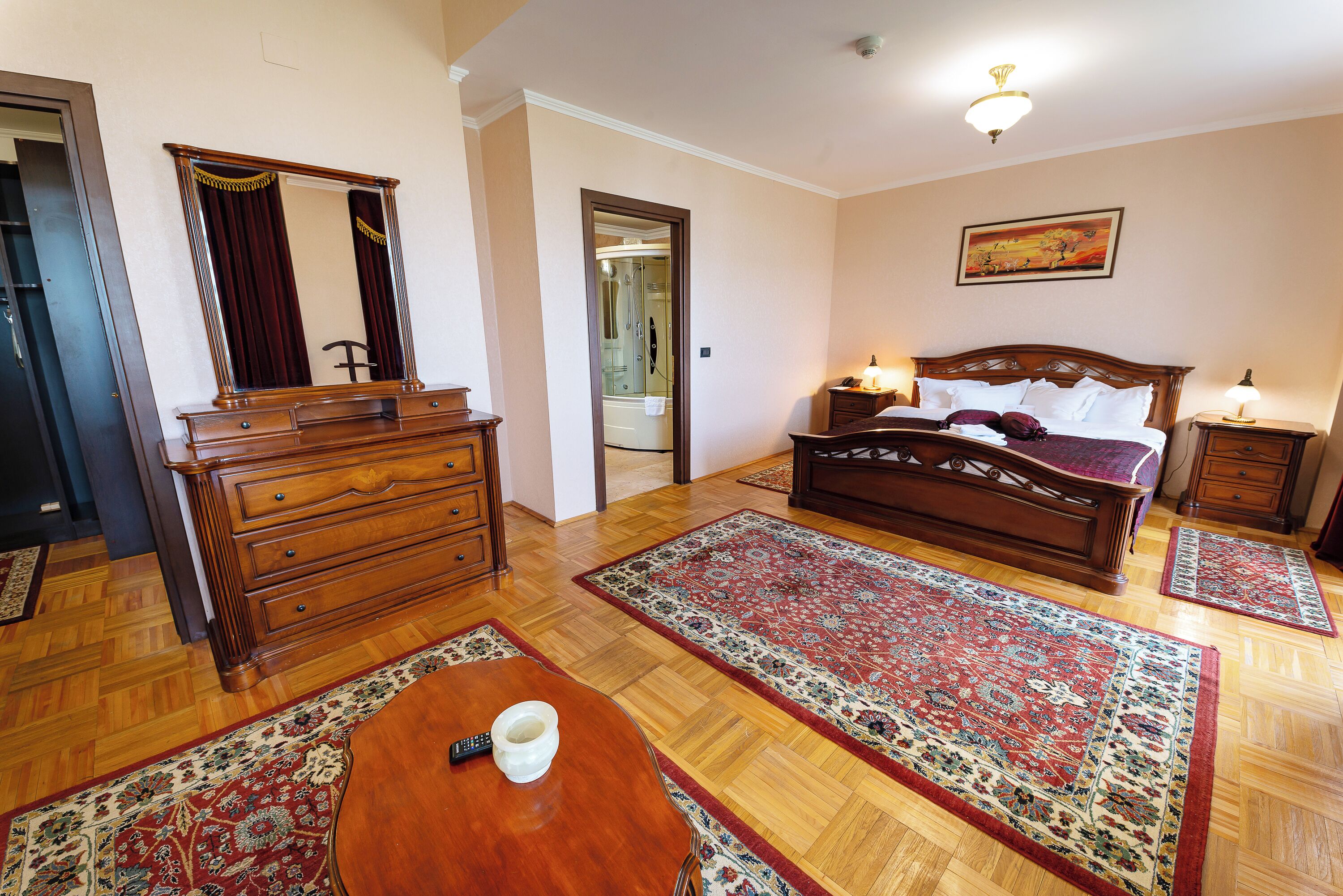 Quarto de hotel com cômoda de madeira, cama com cabeceira, mesa de cabeceira, escrivaninha e um tapete grande.
