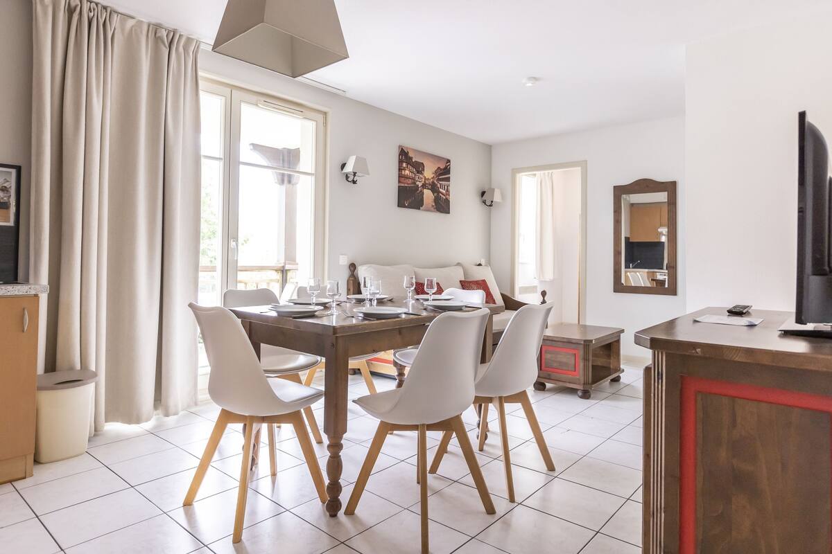 Uma área de jantar moderna com mesa de madeira, cadeiras brancas e uma grande janela com cortinas.
