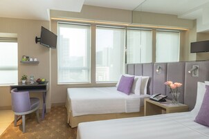 Quarto de hotel com duas camas, uma escrivaninha com cadeira, televisão na parede e vista para a cidade através de grandes janelas.