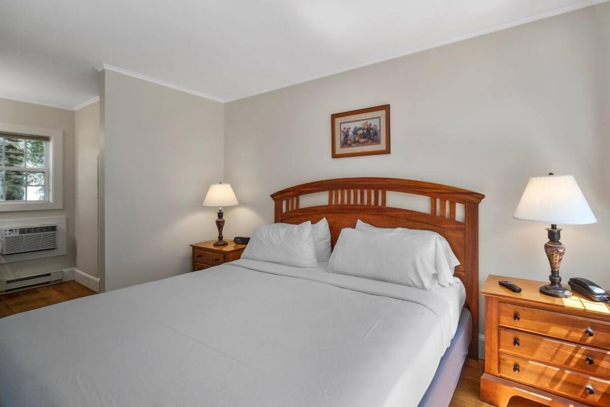 Uma cama bem arrumada com lençóis brancos, duas mesas de cabeceira com abajures, uma cabeceira de madeira e um quadro na parede.