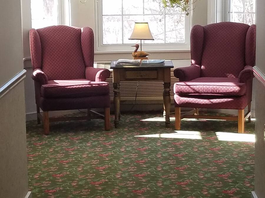 Um quarto com duas poltronas vermelhas, uma pequena mesa de madeira com abajur e um tapete estampado com flores.