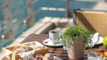 Mesa posta com doces, acompanhamentos e uma planta em vaso em um deck com guarda-corpo e vista para o mar.