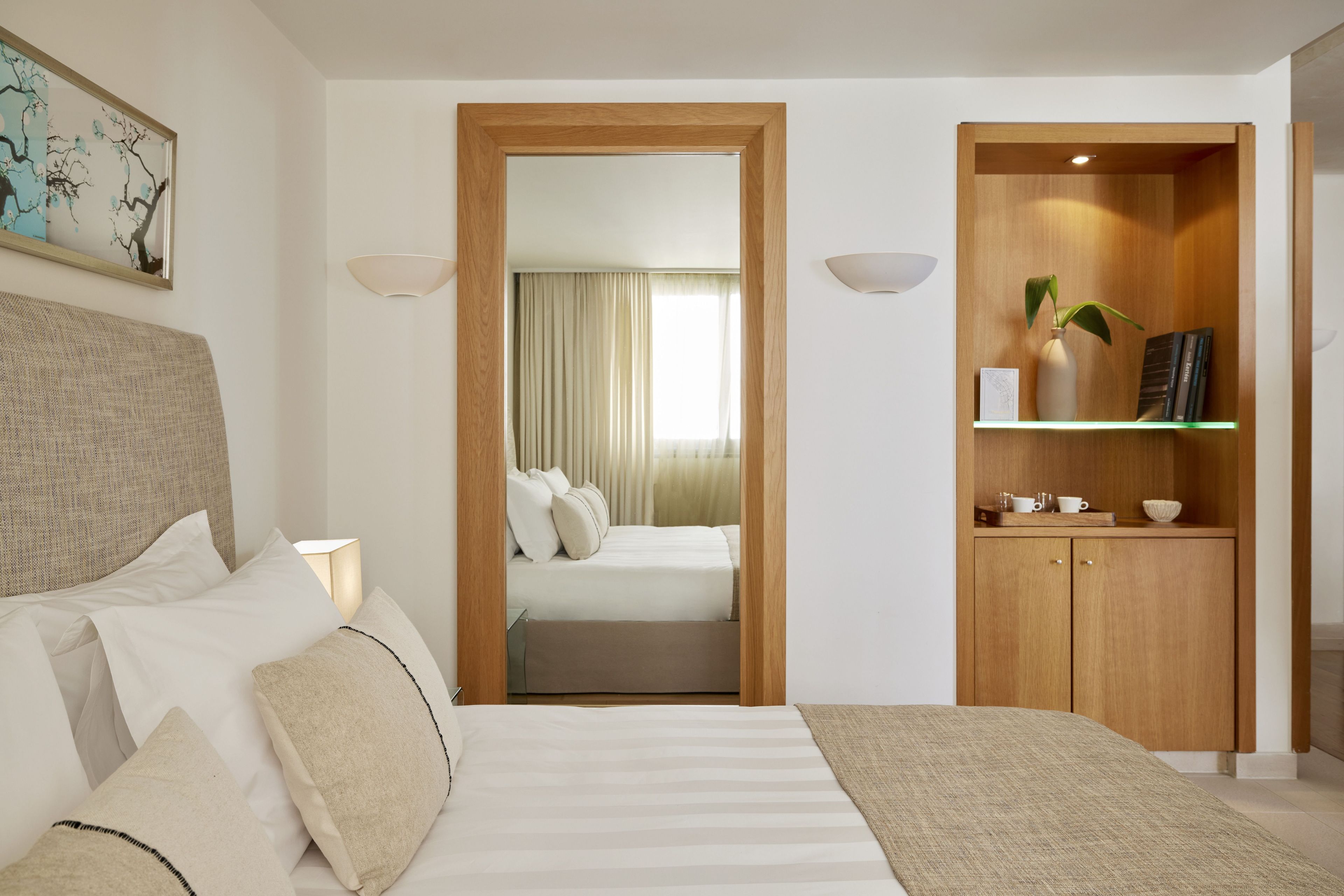 Quarto de hotel com uma cama, abajures, um armário de madeira com uma planta e um quadro na parede.