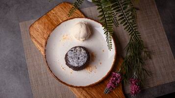 Prato de sobremesa com bolo de chocolate e sorvete sobre uma tábua de madeira, acompanhado de samambaia e flores rosa.