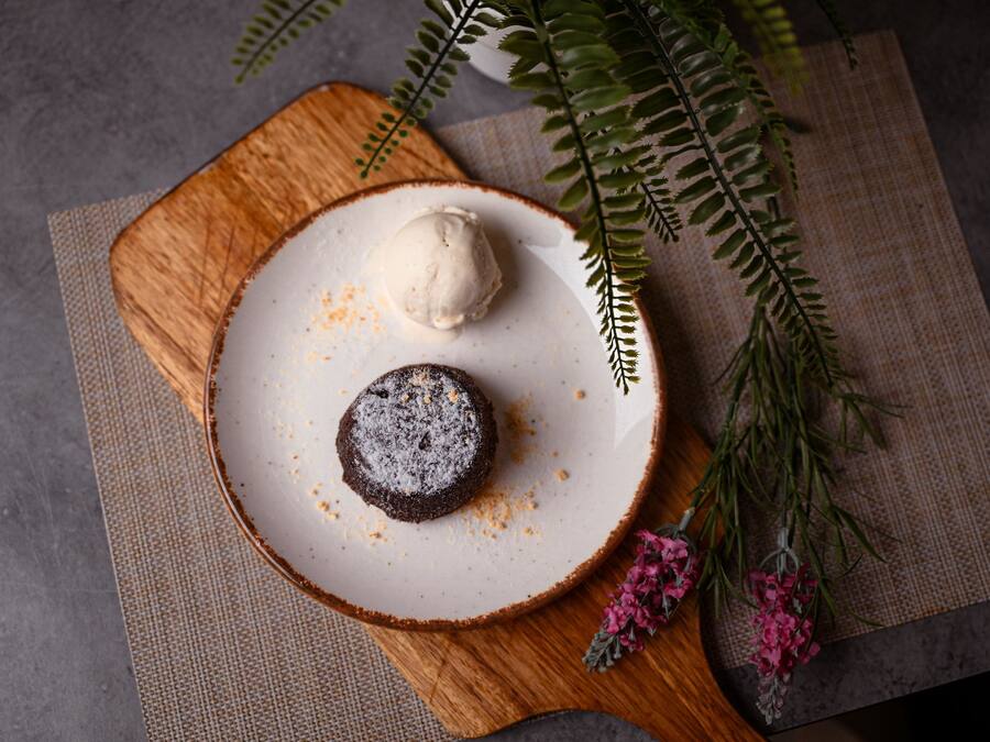 Prato de sobremesa com bolo de chocolate e sorvete sobre uma tábua de madeira, acompanhado de samambaia e flores rosa.