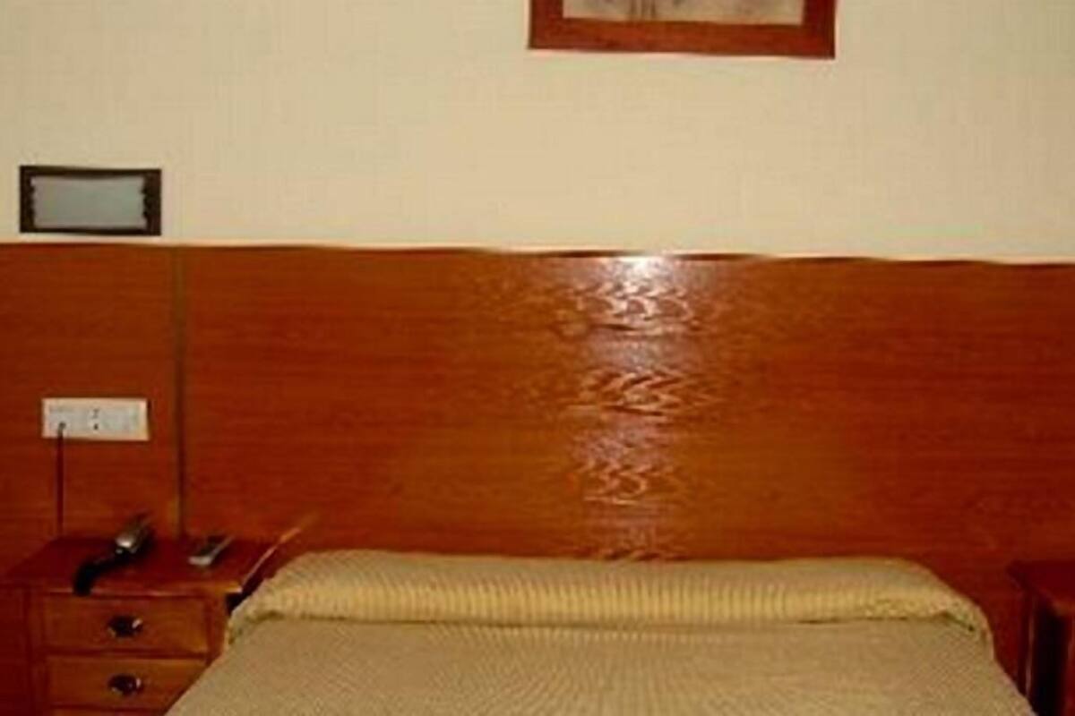 Quarto de hotel com cabeceira de madeira, cama com manta bege, duas mesas de cabeceira e duas luminárias de parede.