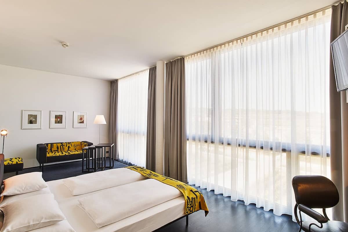 Quarto de hotel com cama, escrivaninha, cadeira e janelas amplas com cortinas leves.