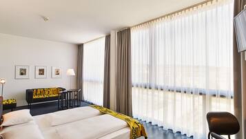 Quarto de hotel com cama, escrivaninha, cadeira e janelas amplas com cortinas leves.