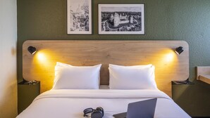 Cama bem arrumada com lençóis brancos, um laptop e fones de ouvido sobre a cama. Há duas quadros na parede acima da cama.
