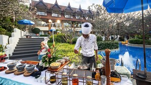 Um cozinheiro prepara alimentos ao ar livre, com uma piscina e um edifício tradicional ao fundo.