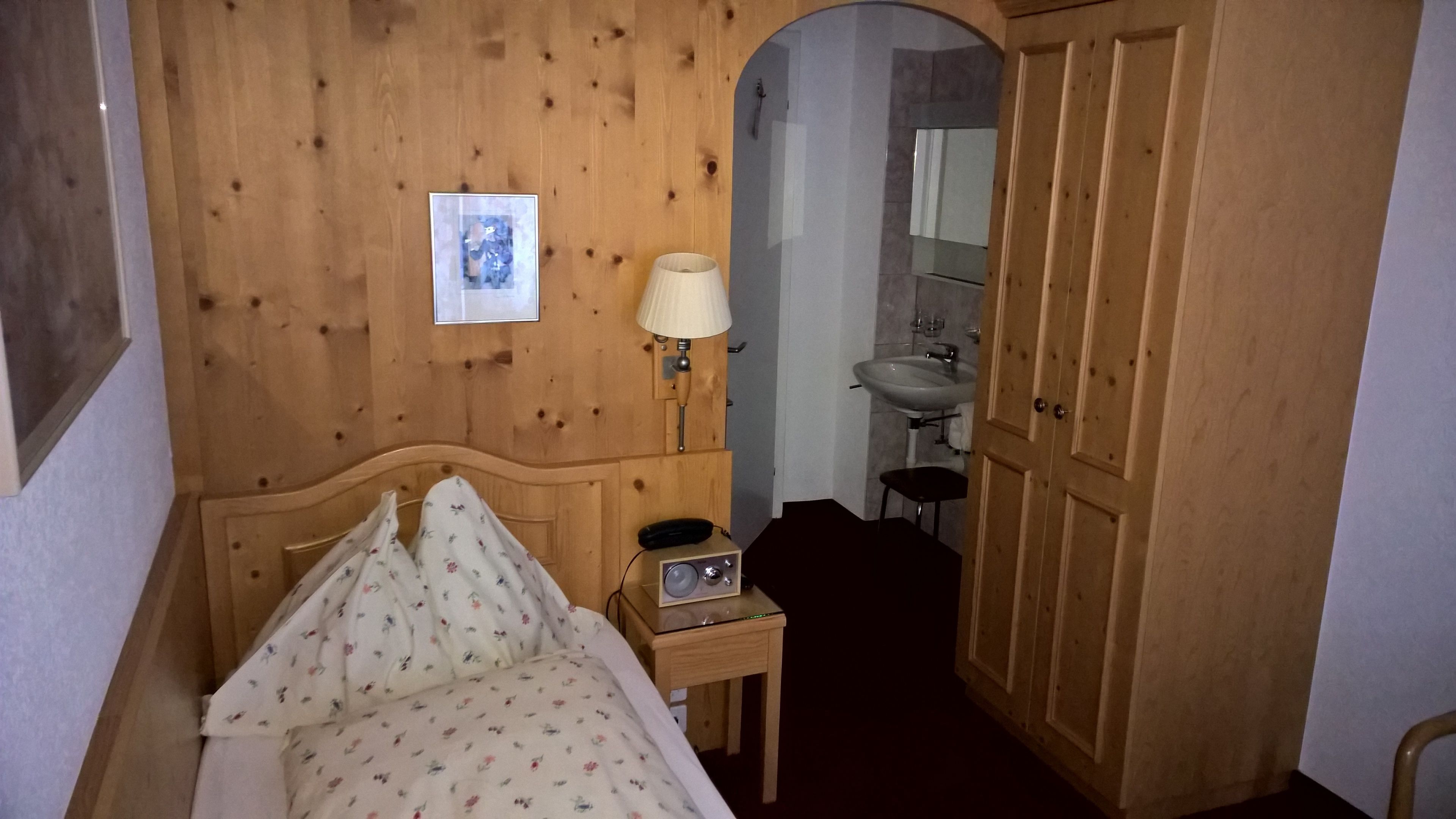 Quarto com painéis de madeira, com uma cama, mesa de cabeceira com abajur, pequena mesa com rádio e um banheiro visível por um arco.