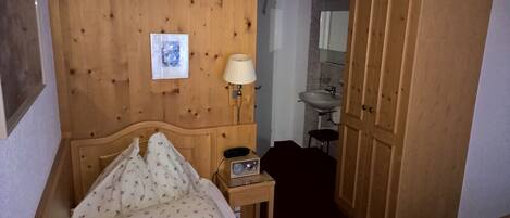 Quarto com painéis de madeira, com uma cama, mesa de cabeceira com abajur, pequena mesa com rádio e um banheiro visível por um arco.