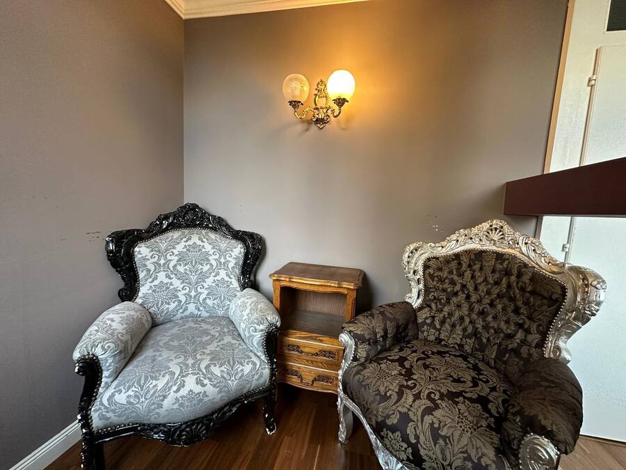 Quarto de canto com duas cadeiras elaboradas, uma pequena mesa lateral de madeira e um aplique de parede.