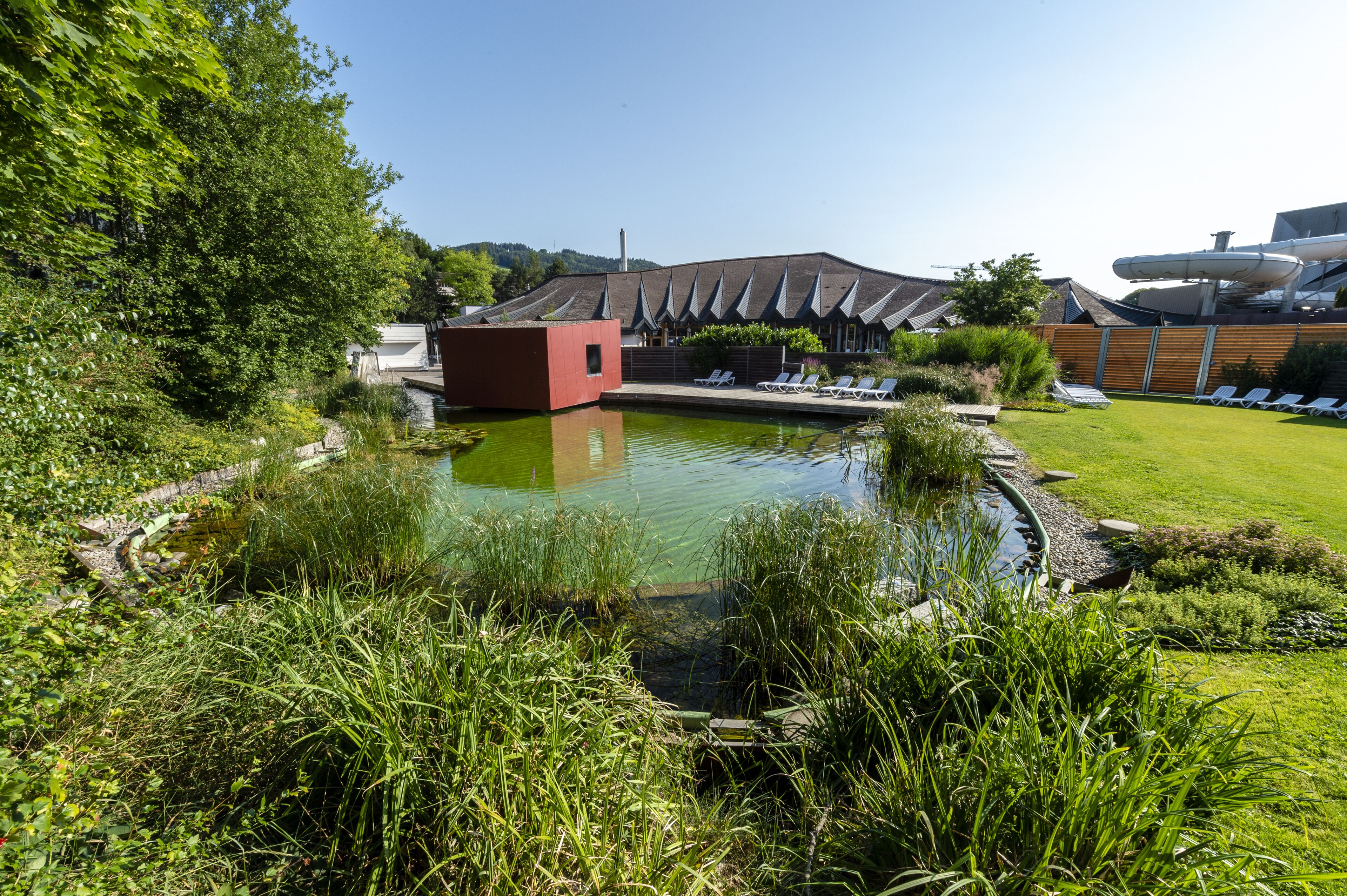 Piscina rodeada por vegetação exuberante, um edifício vermelho e uma estrutura moderna com telhado inclinado.