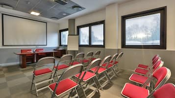Uma sala de conferências com fileiras de cadeiras dobráveis, uma tela de projetor e janelas com persianas.