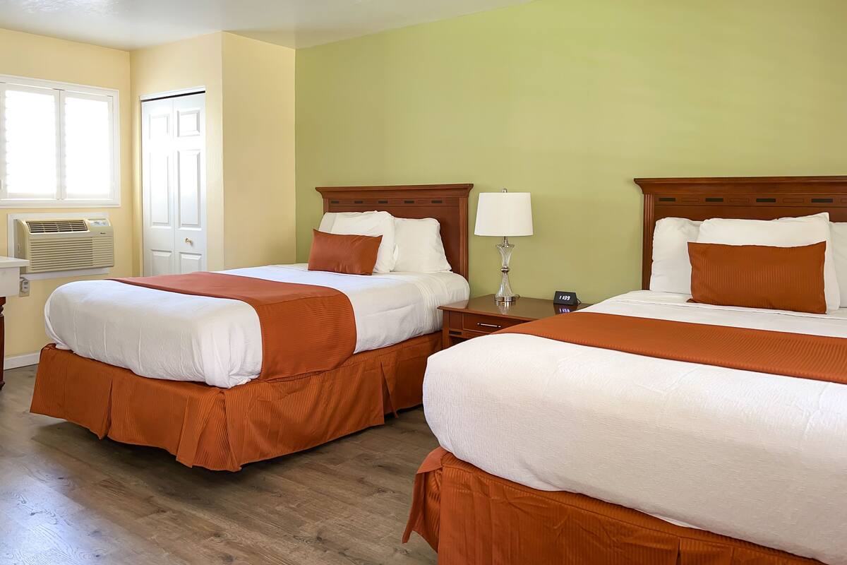 Quarto de hotel com duas camas, cada uma com roupa de cama branca e detalhes em laranja, piso de madeira, janela com persianas e ar-condicionado fixado na parede.
