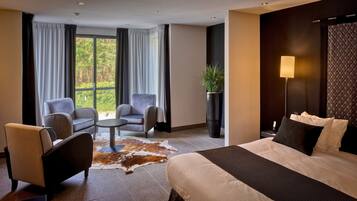 Quarto de hotel moderno com uma cama grande, duas poltronas, uma mesa pequena e vista para o exterior através de amplas janelas.