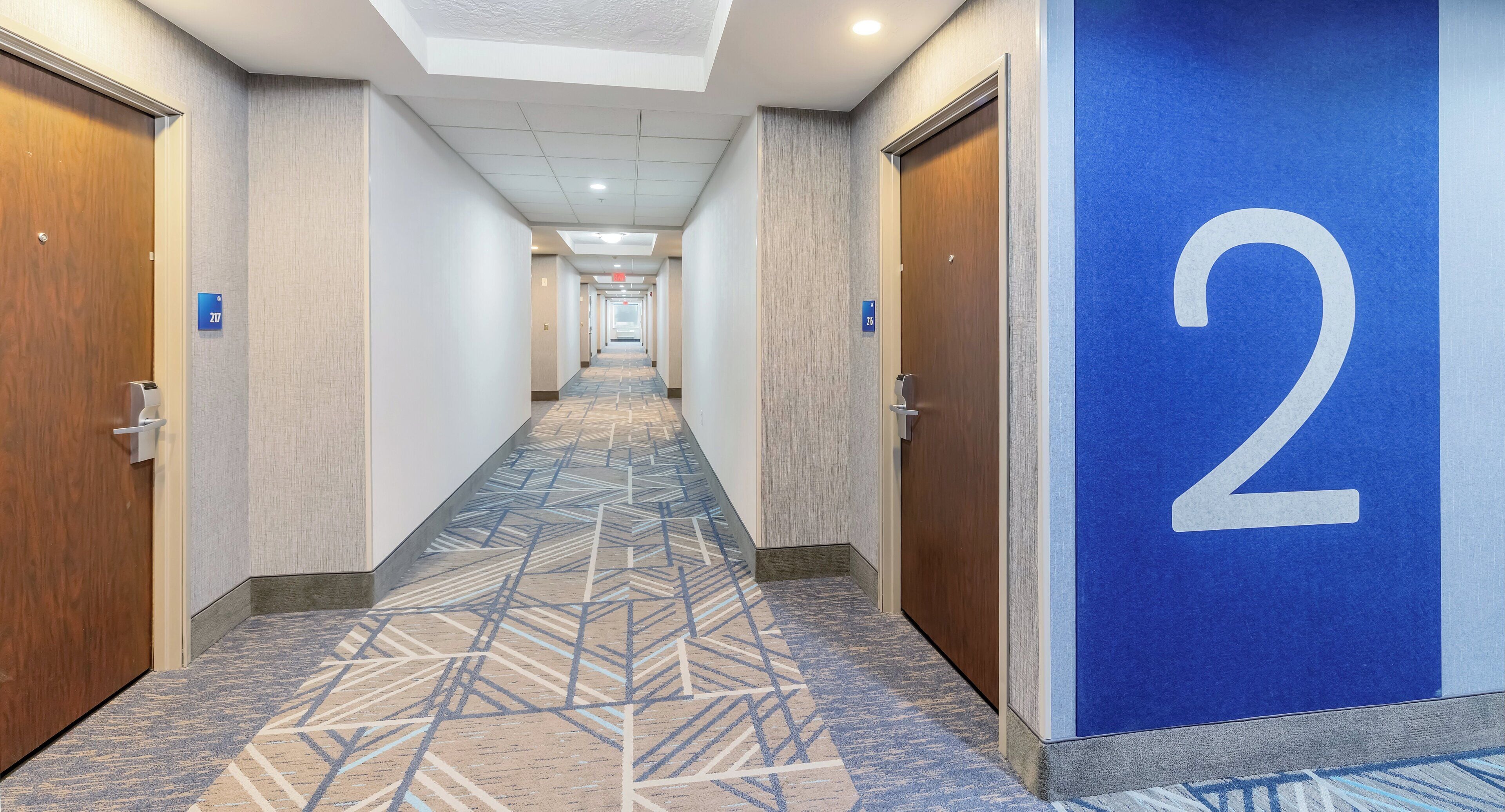 Um corredor de hotel com carpete estampado, portas de madeira e parede azul com o número 2.