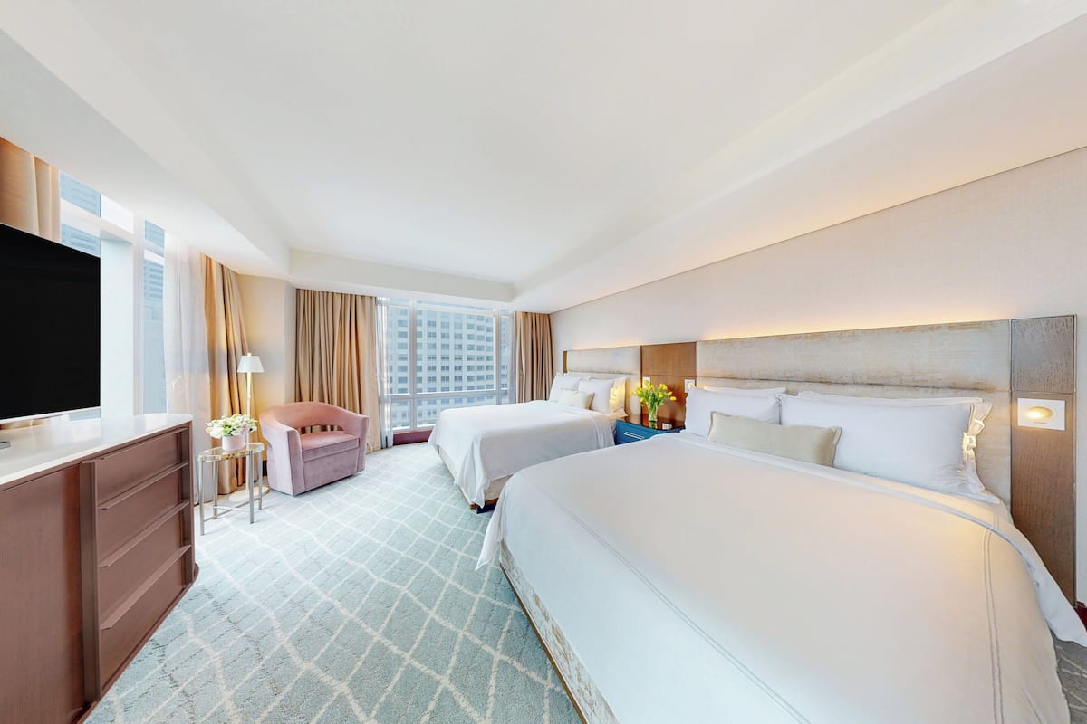 Quarto de hotel com duas camas, uma mesa, uma cadeira, televisão e uma janela grande.