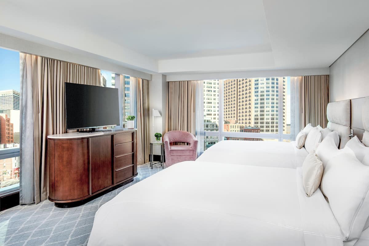 Quarto de hotel com uma cama grande, televisão sobre um armário de madeira, uma cadeira rosa e vista para a cidade.