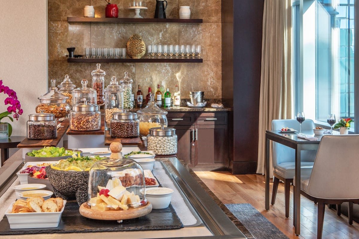 Disposição de buffet com diversos alimentos e acompanhamentos, área de estar com mesa e cadeiras, e uma grande janela que oferece luz natural.
