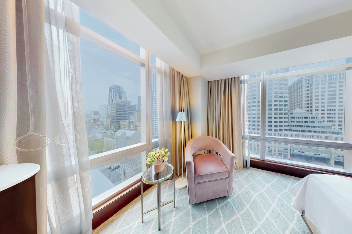 Quarto de hotel com uma cama, uma cadeira, uma mesinha com um vaso de flores e janelas amplas com vista para a cidade.