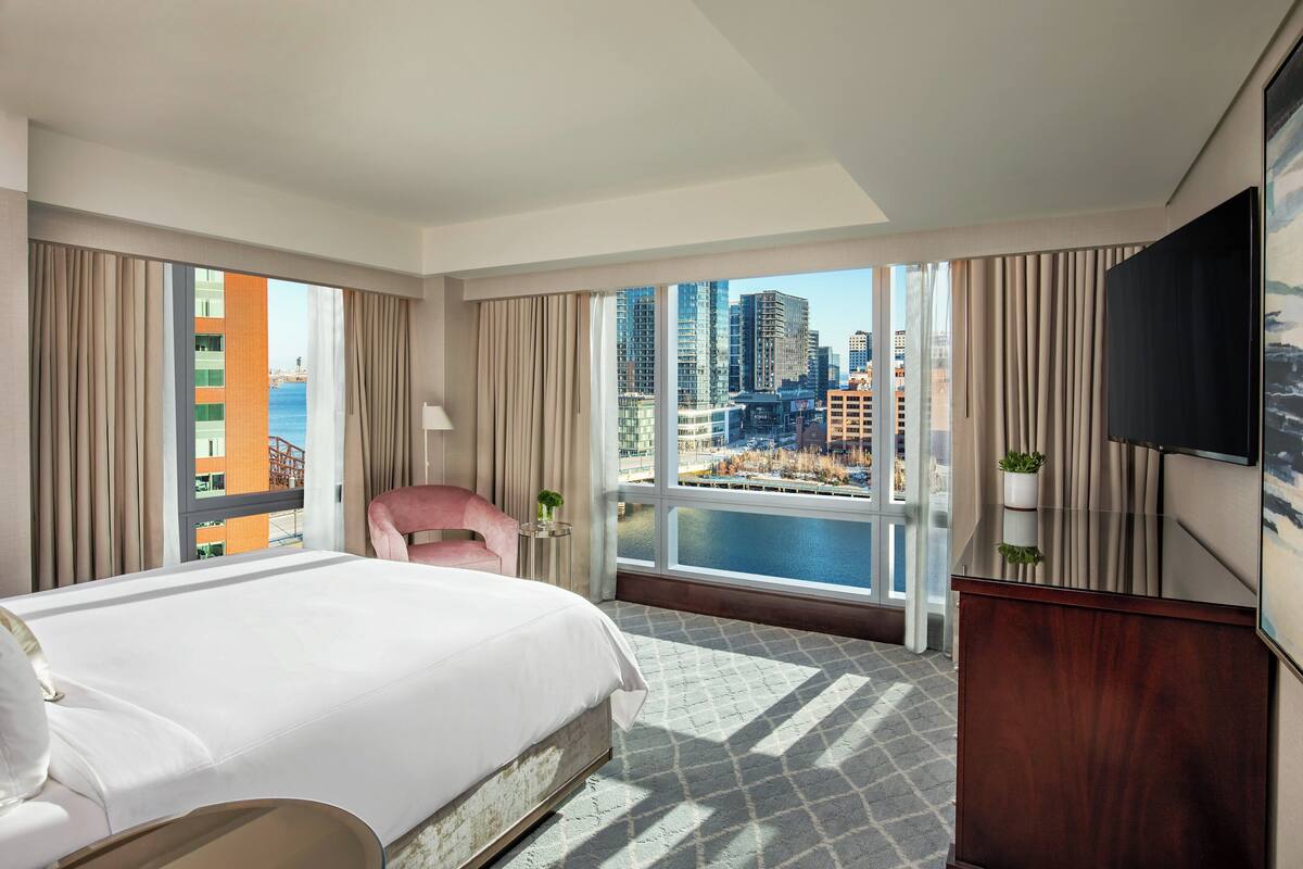 Quarto de hotel com uma cama grande, televisão, escrivaninha e vista para a paisagem urbana através de amplas janelas.