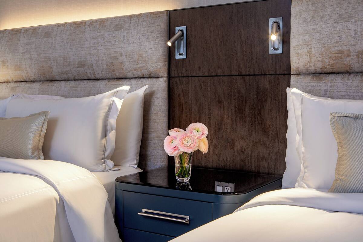 Quarto de hotel com cabeceira de madeira, mesa de cabeceira com um vaso de flores e um criado-mudo com gaveta.