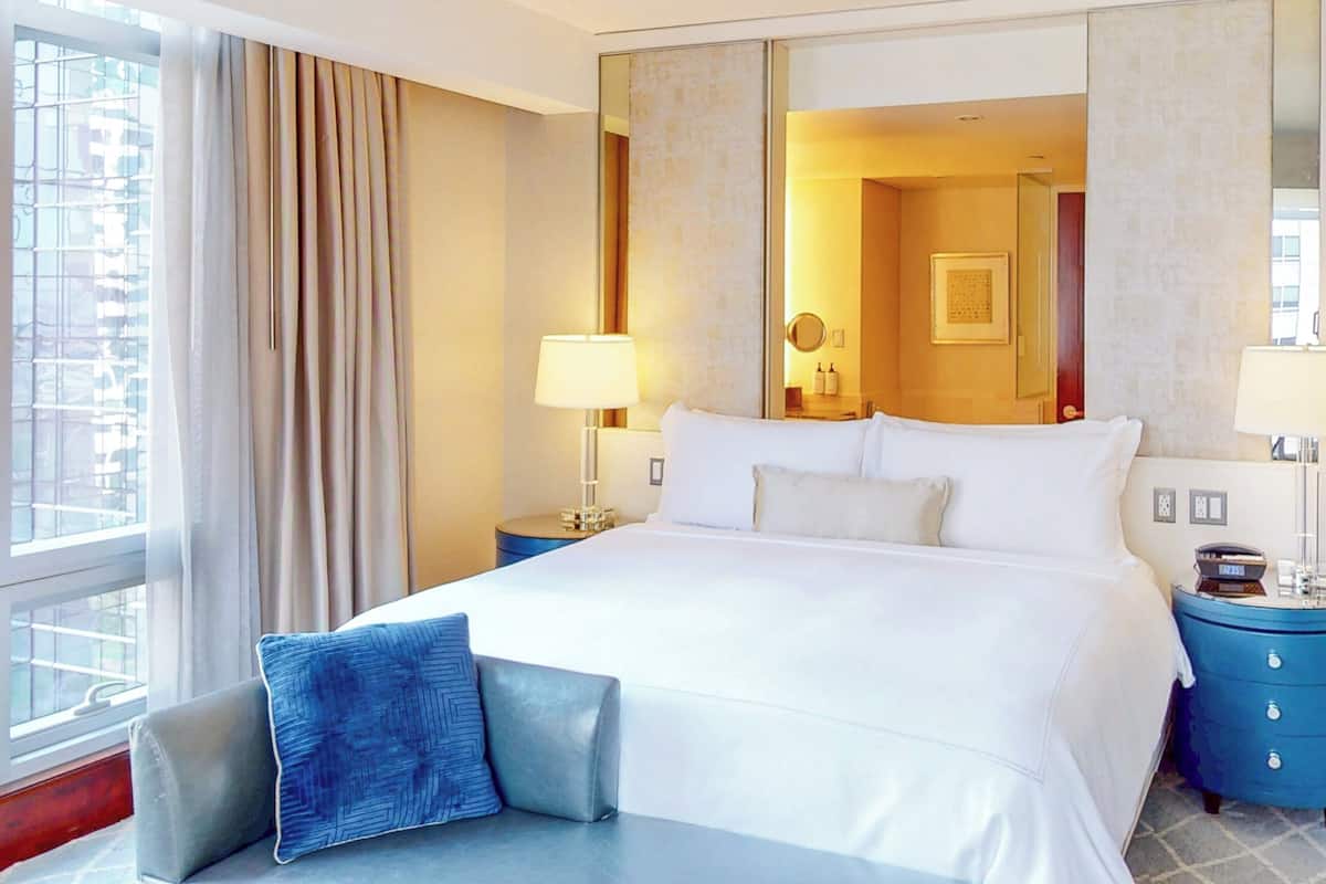 Quarto de hotel com uma cama grande, duas mesas de cabeceira, uma cadeira azul e vista para a cidade pela janela.