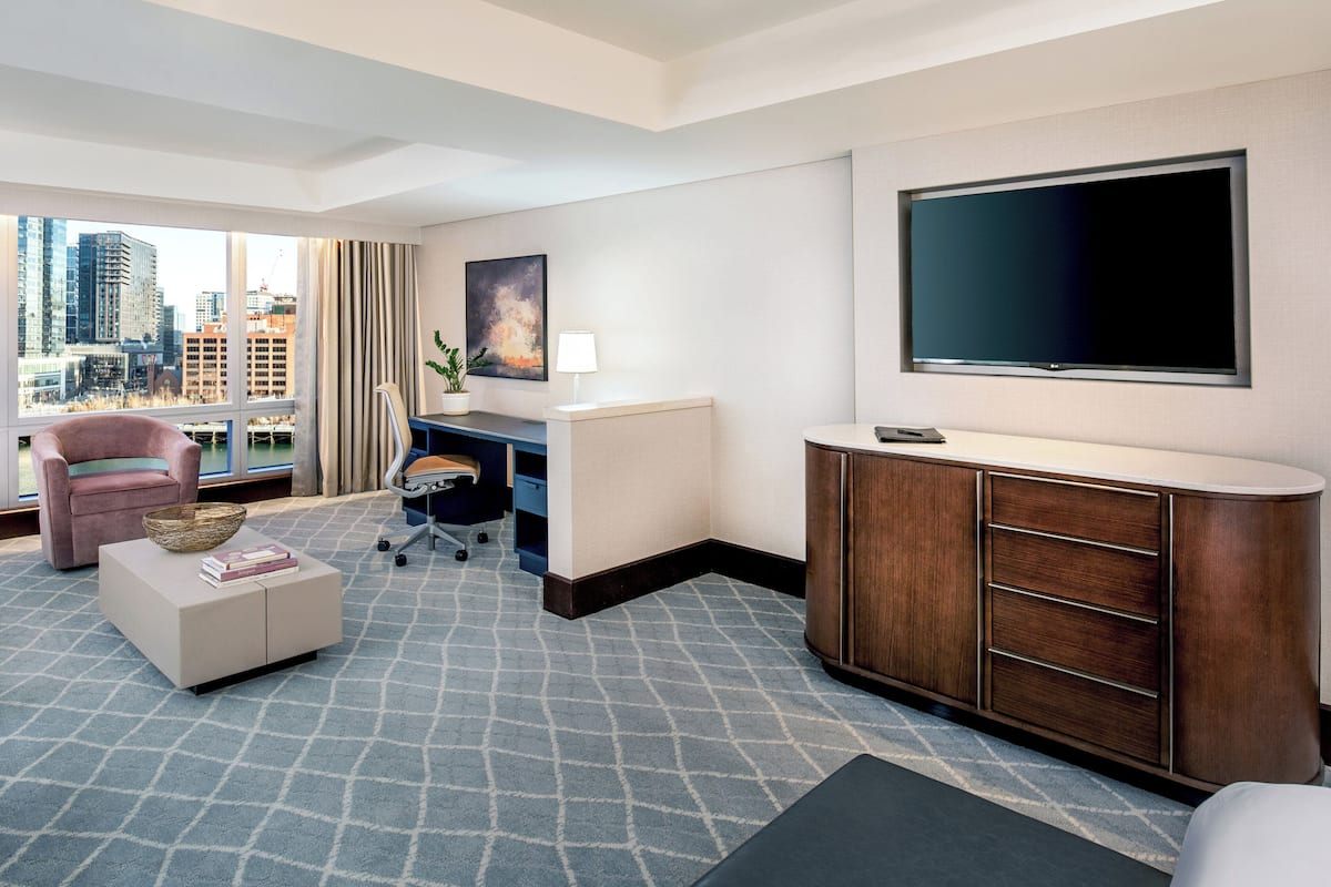 Quarto de hotel com uma janela grande, televisão de tela plana, escrivaninha com abajur, cadeira, sofá e armário.