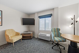 Quarto de hotel com televisão de tela plana, sofá, poltrona estampada, mesa pequena e abajur.