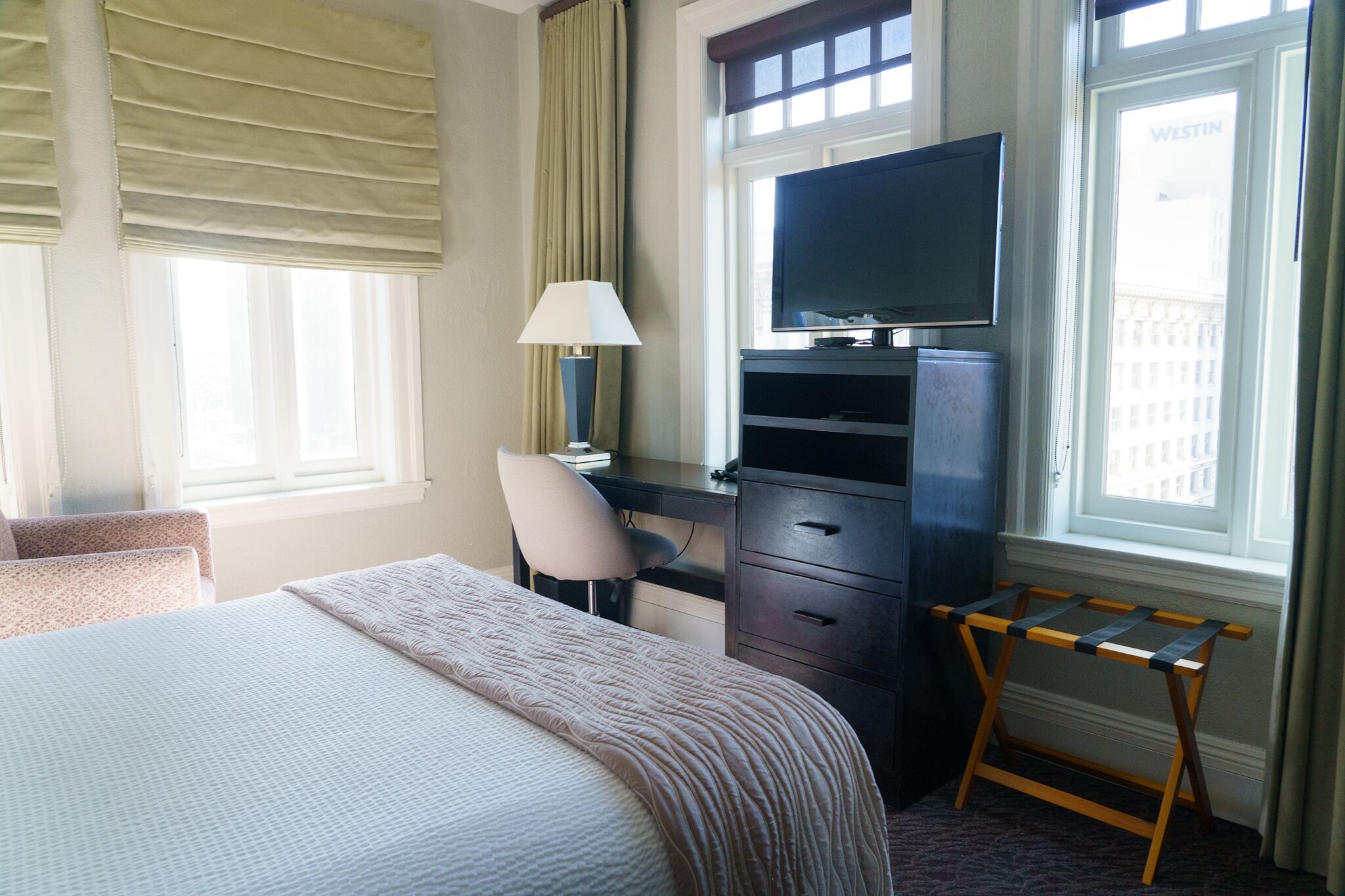 Quarto de hotel com cama, escrivaninha com televisão, cadeira e janela com cortinas.