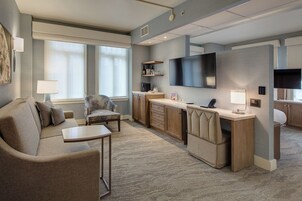 Quarto de hotel moderno com sofá, poltrona, escrivaninha e televisão de tela plana.
