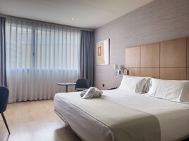 Quarto de hotel com cama grande, mesa pequena, cadeira, luminária na parede e janela com cortinas.