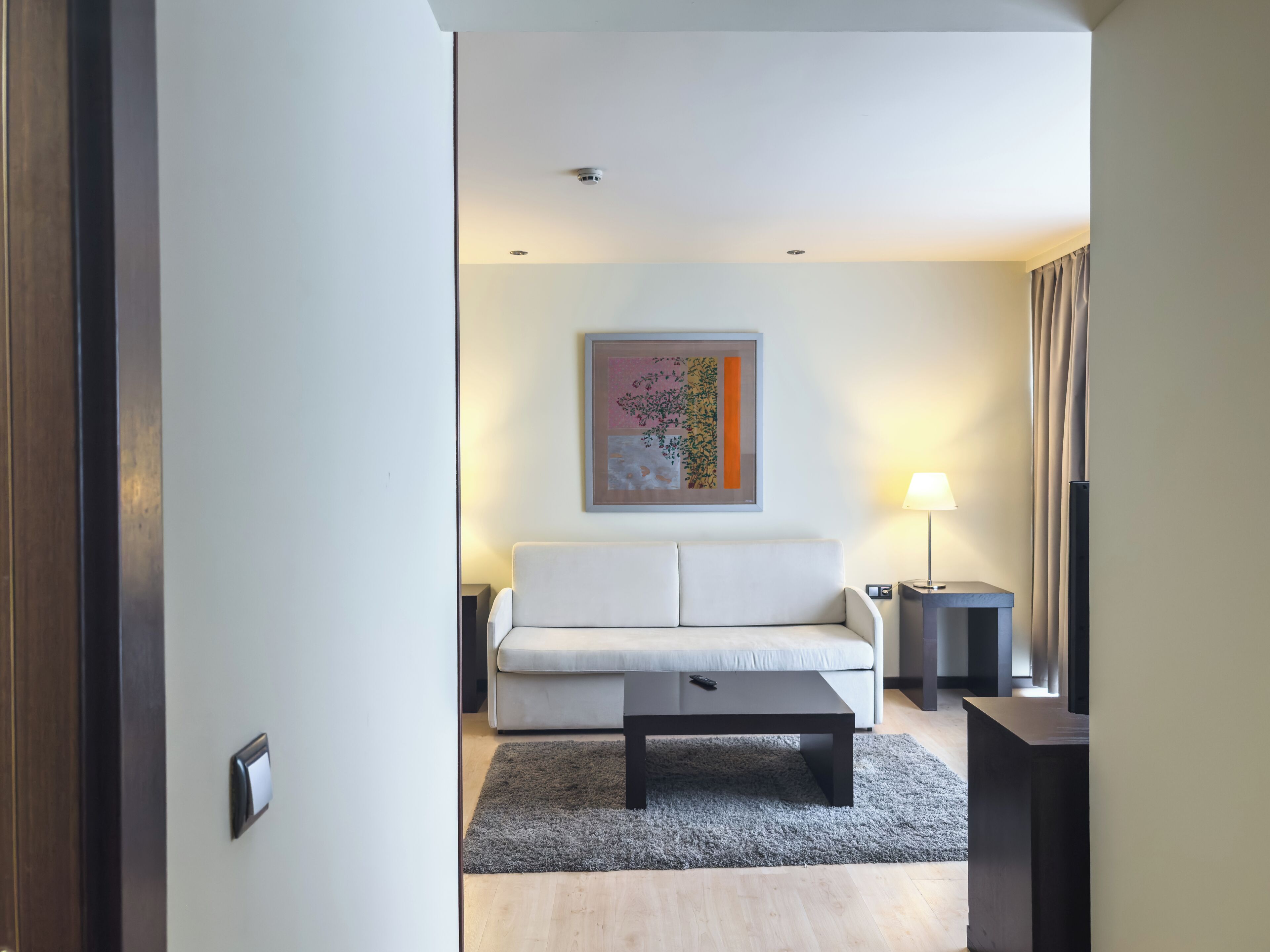 Uma sala de estar moderna com um sofá branco, uma mesa de centro preta, um tapete cinza, uma obra de arte emoldurada na parede e um abajur sobre uma mesinha lateral.