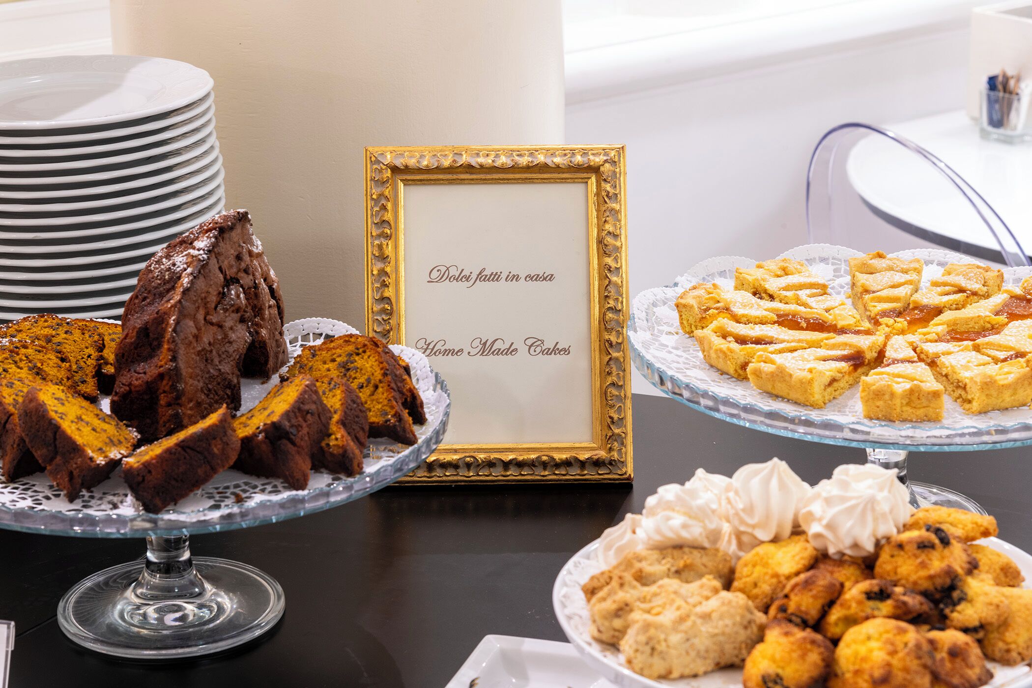 Uma mesa com diversos doces, incluindo bolos e docinhos, uma pilha de pratos e um porta-retrato decorativo com texto.