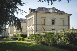 Um edifício grande e histórico, com arquitetura simétrica, vários janelões e um jardim bem cuidado.