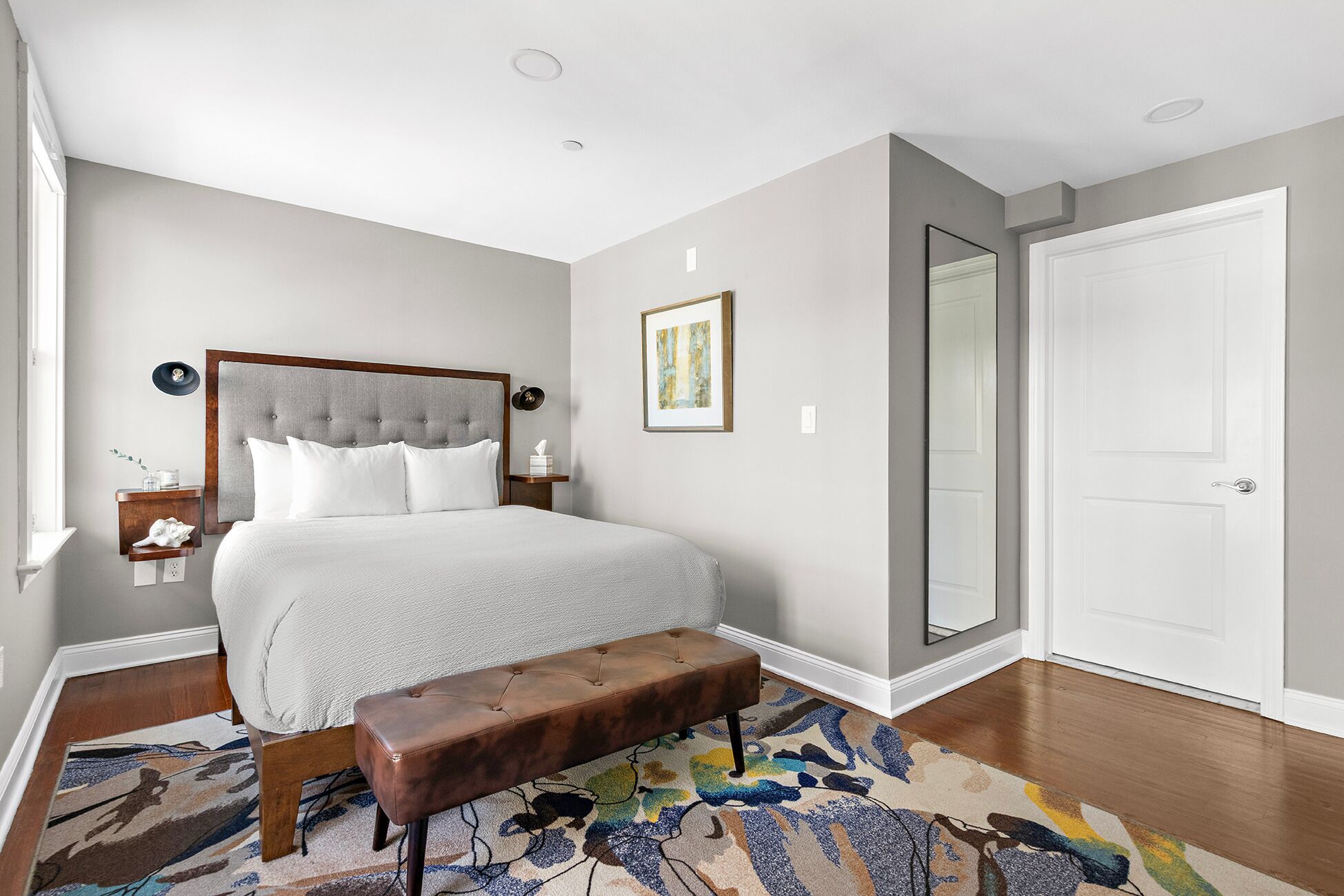 Quarto com cama, banqueta, mesa de cabeceira e porta.