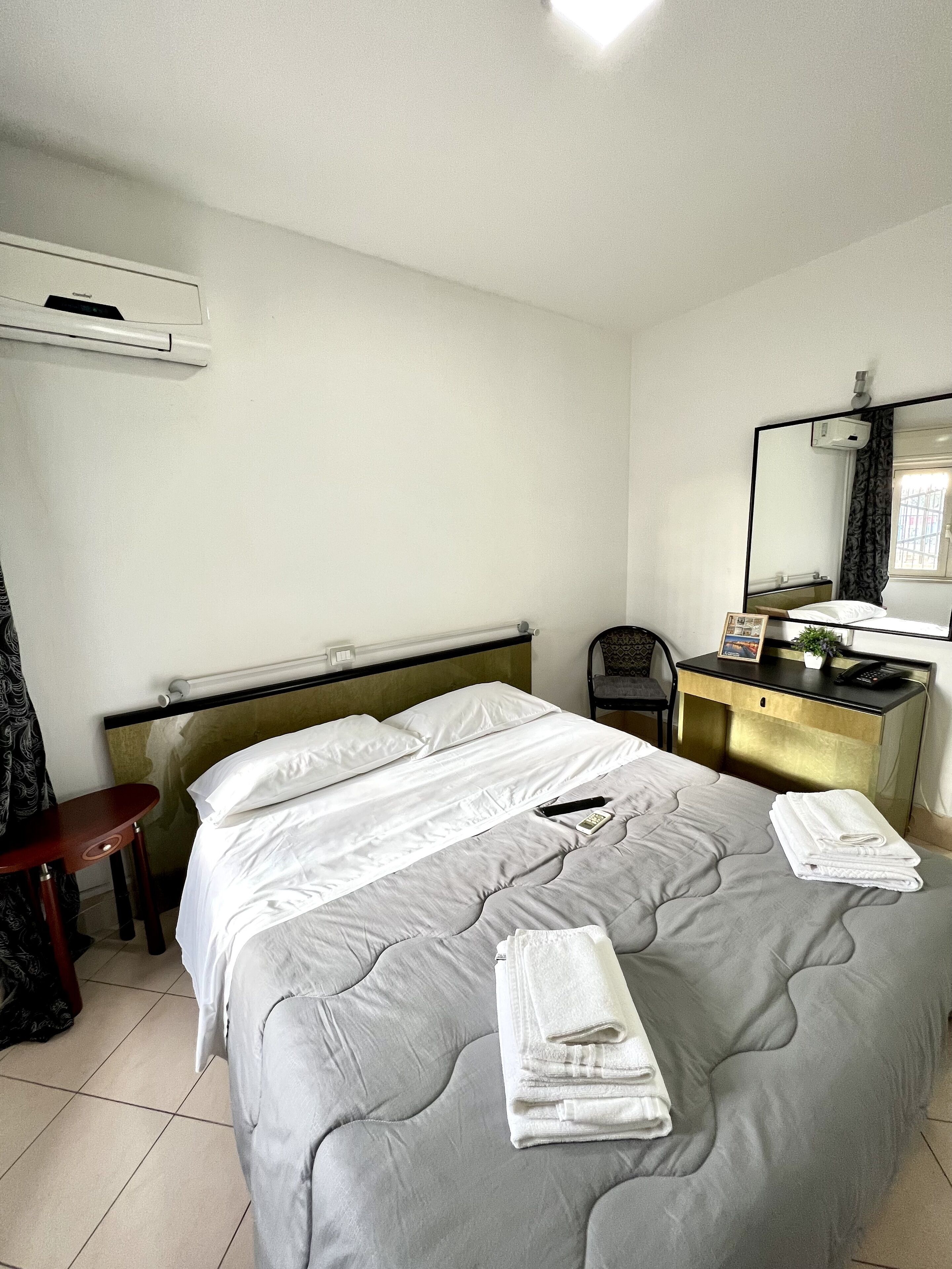 Quarto de hotel com cama, mesa de cabeceira, cadeira, escrivaninha e espelho.