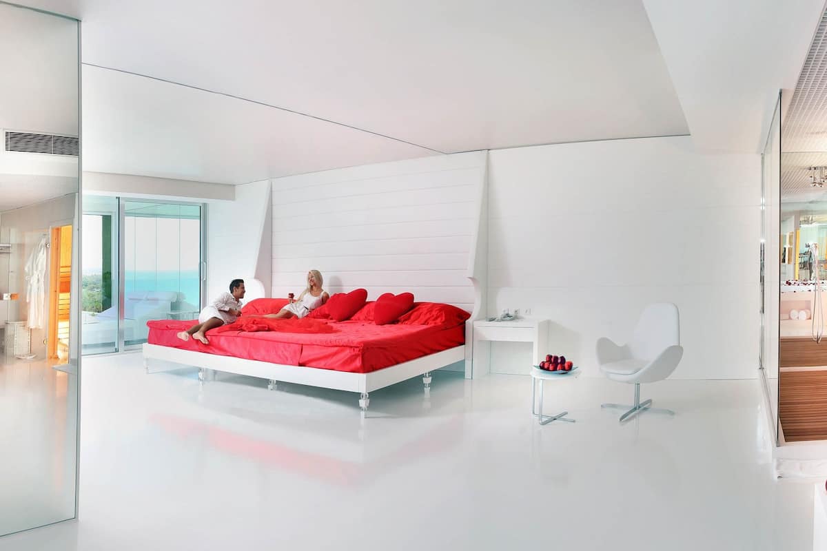 Quarto de hotel moderno com uma cama grande, parede de vidro e vista para a cidade.