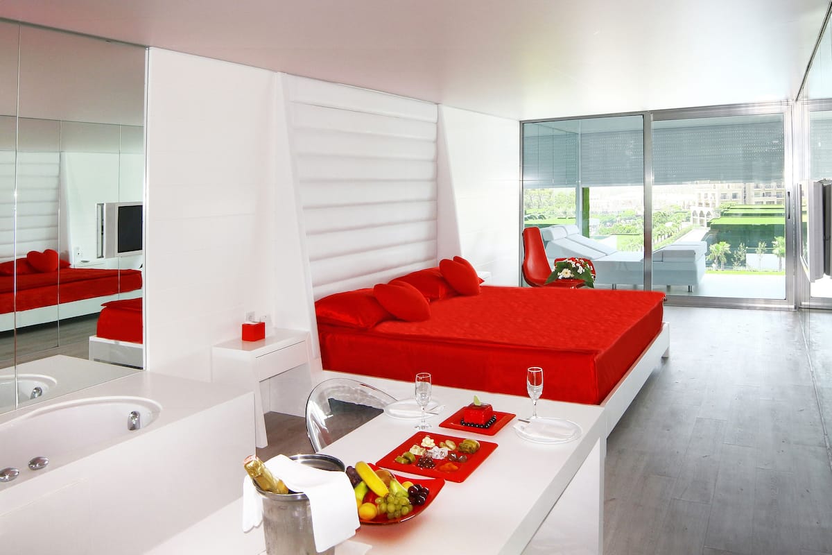 Quarto de hotel moderno com um sofá vermelho, um espelho grande, uma mesa de cabeceira branca com uma garrafa de champanhe e vista para uma paisagem verde através de grandes janelas.