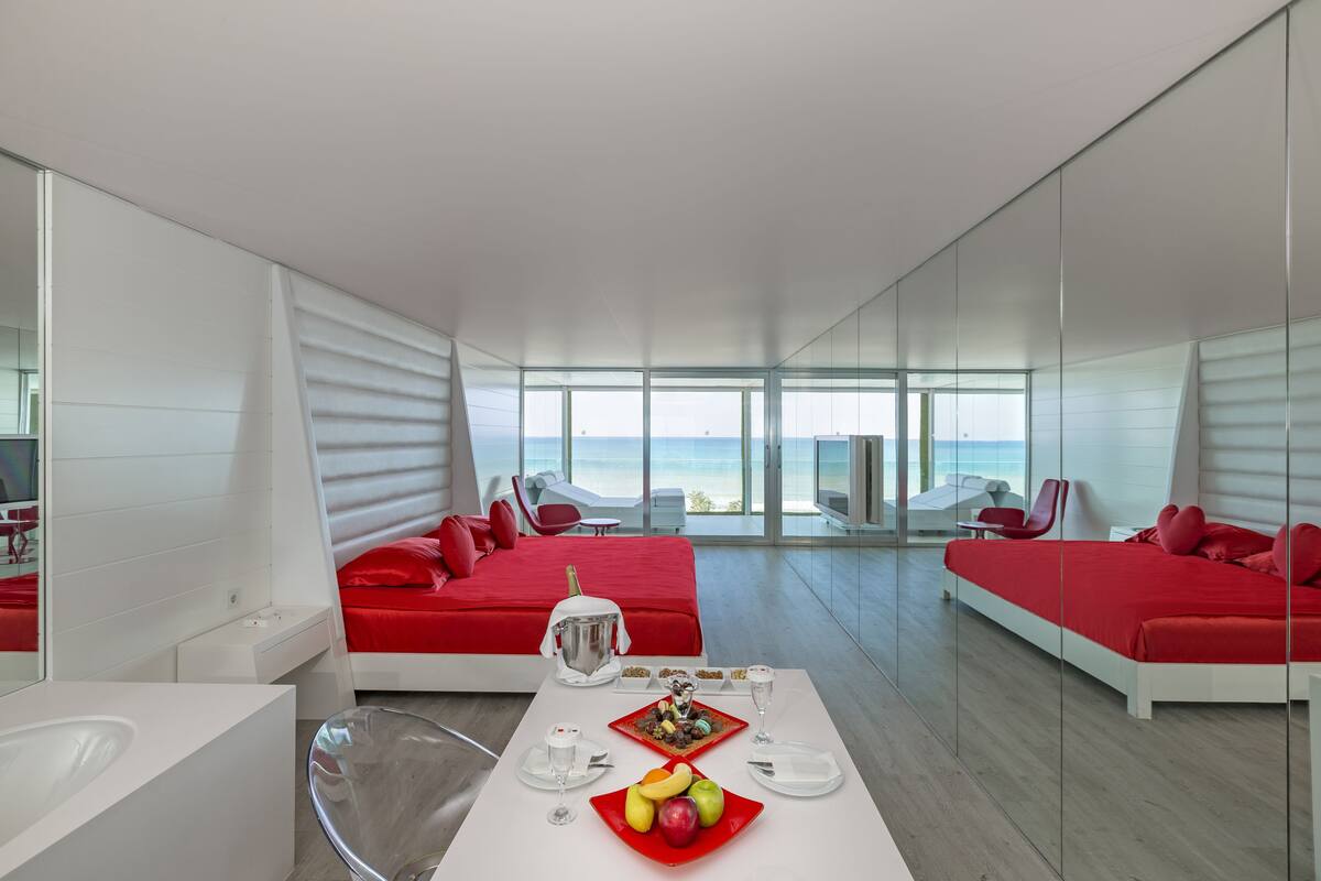 Quarto de hotel moderno com uma cama grande, uma mesa de jantar com frutas e bebidas, e vista para o oceano.