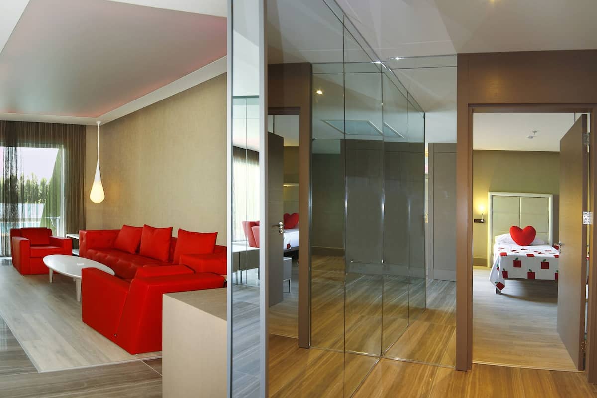 Quarto de hotel moderno com sofá vermelho, mesa de centro branca, TV e divisória de vidro.