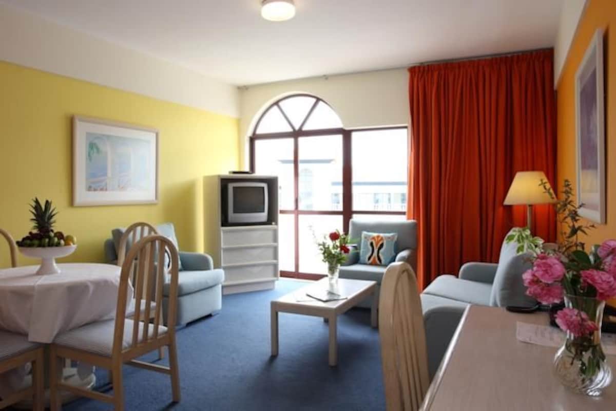 Uma sala de estar com uma mesa de jantar, dois sofás, uma televisão e uma janela com cortinas.
