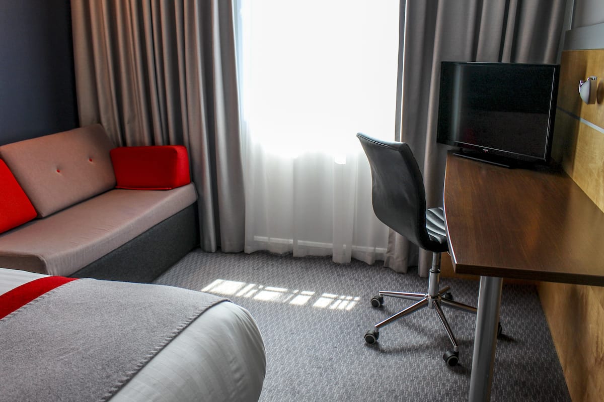 Quarto de hotel com cama, escrivaninha com televisão, poltrona e sofá com almofadas vermelhas.