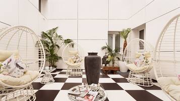 Um hall amplo com piso xadrez, cadeiras de vime branco, plantas em vasos e um vaso central.