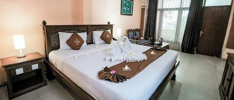 Quarto de hotel com uma cama grande, mesa de cabeceira com abajur, mesa lateral e uma janela com cortinas.