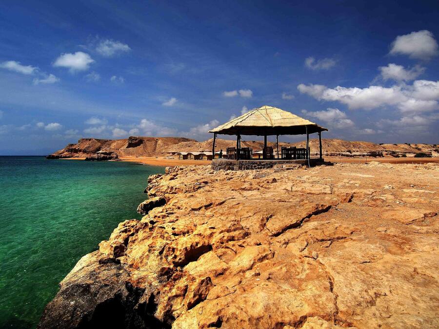 Costa rochosa com uma construção de telhado de palha, água turquesa cristalina e colinas ao longe.